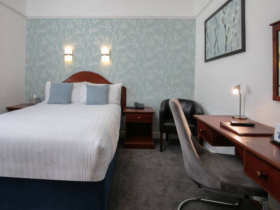 Quarto de hotel com cama, mesa de cabeceira, escrivaninha, cadeira e luminária na parede.