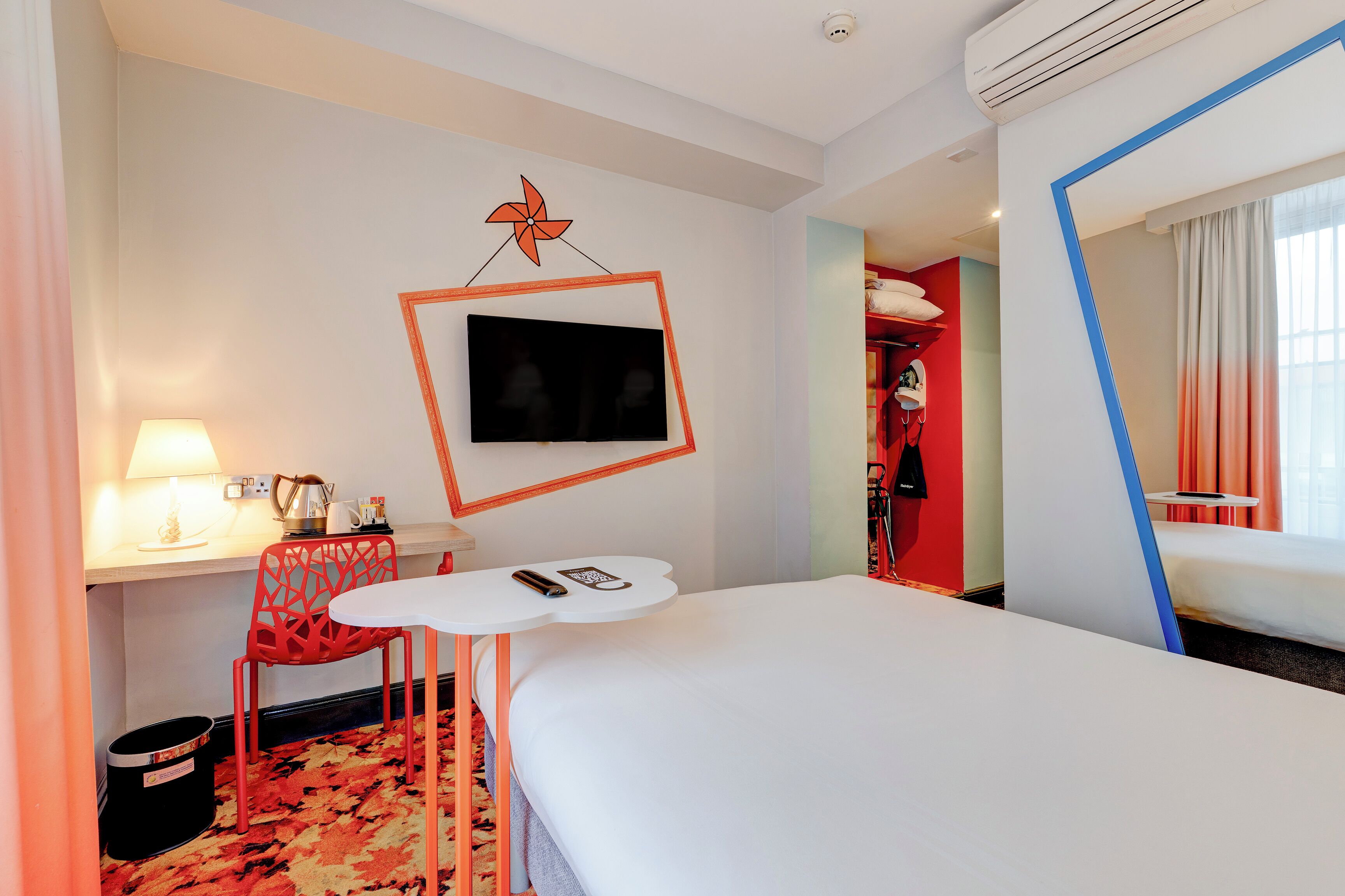 Quarto de hotel com cama, uma mesinha, uma cadeira, televisão e espelho.