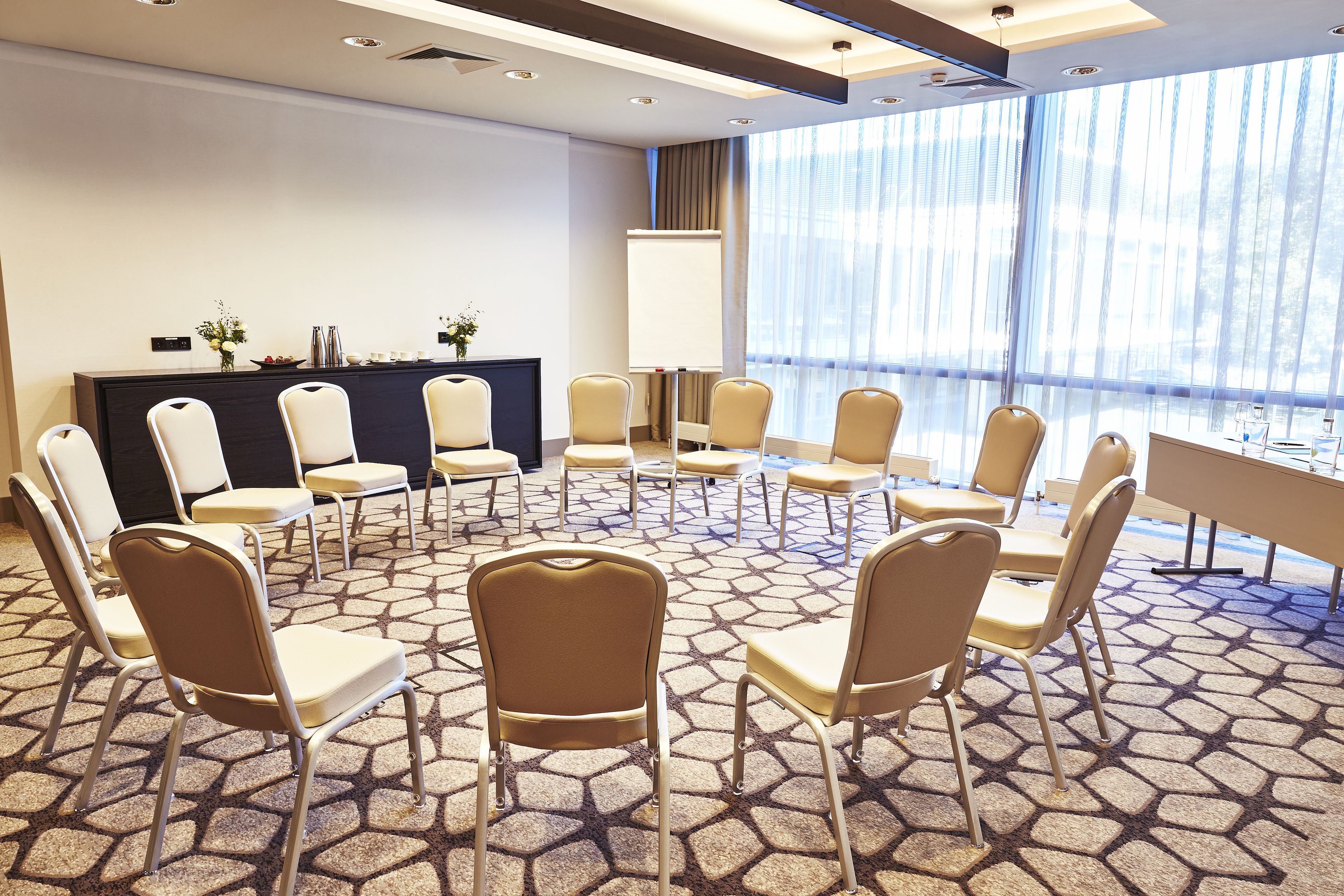 Uma sala de conferências com carpete estampado em padrão hexagonal, cadeiras bege, um balcão de recepção de cor escura e grandes janelas com cortinas leves.