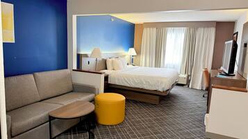 Quarto de hotel com cama, sofá, pequena mesa, um banquinho amarelo, escrivaninha com computador e janela com cortinas.