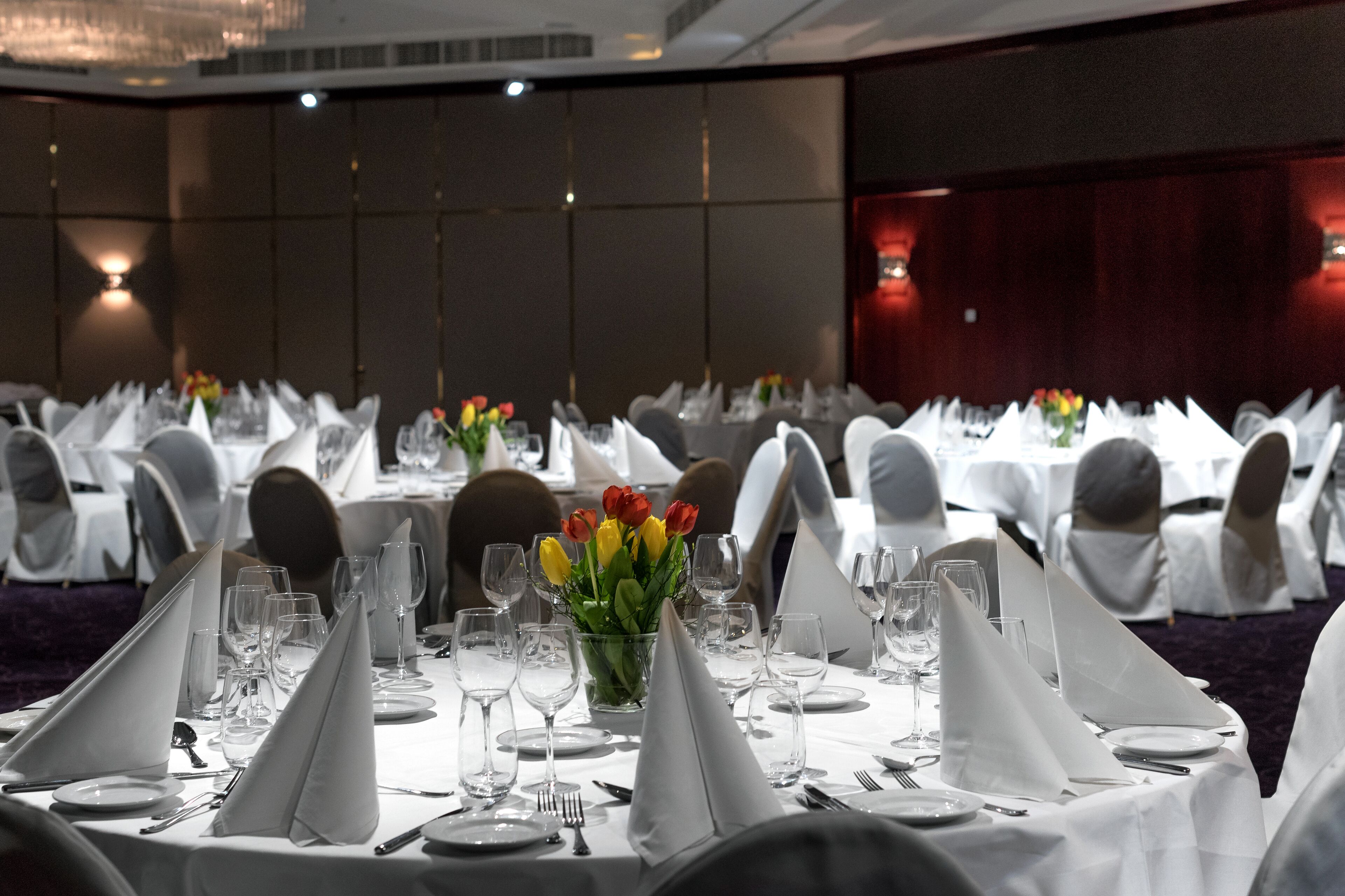 Uma sala de banquetes com mesas montadas para um evento formal, com toalhas brancas, talheres bem dispostos e arranjos florais.