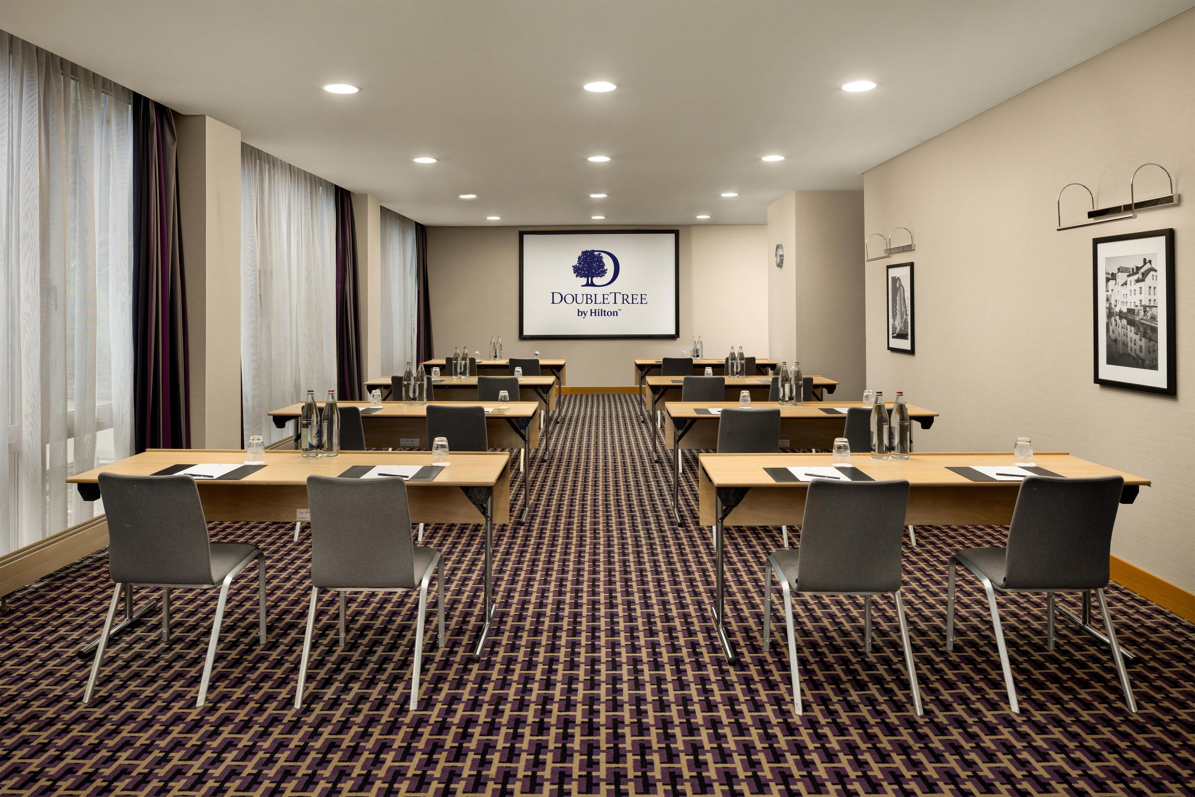 Uma sala de conferências com uma mesa comprida, cadeiras e uma tela exibindo o logotipo da DoubleTree by Hilton.