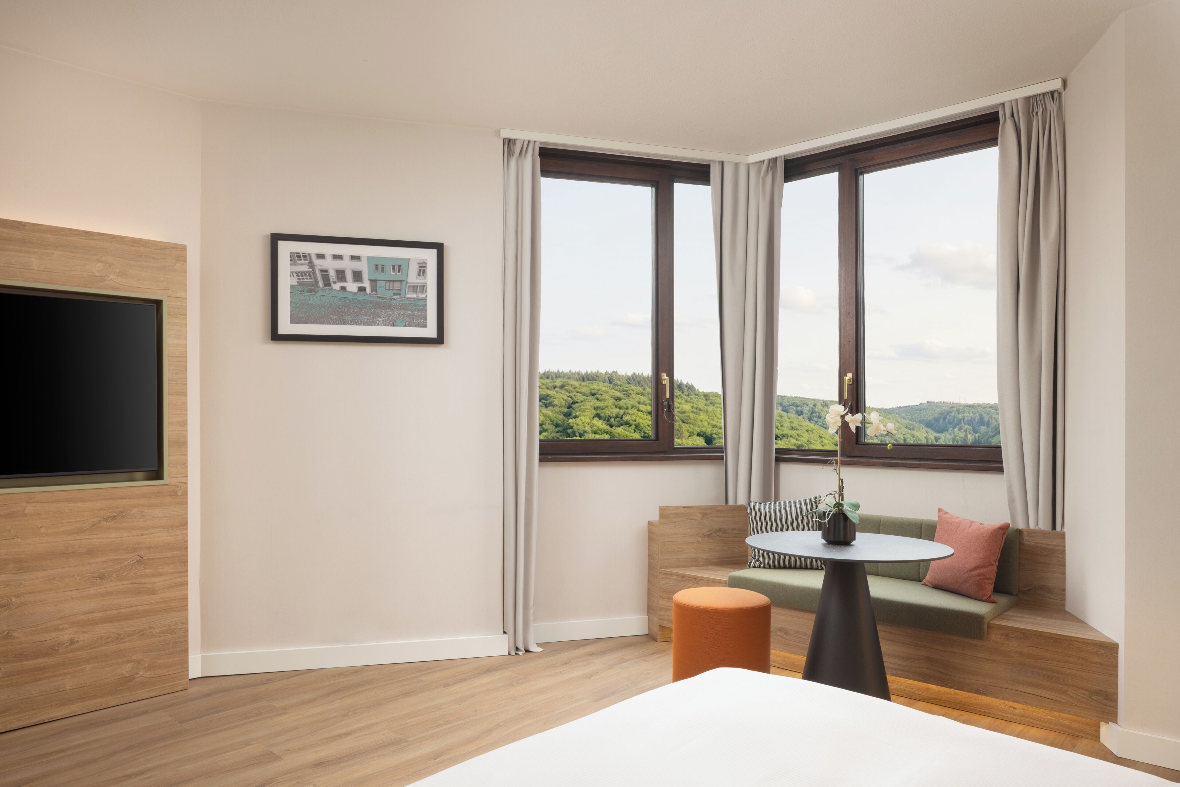 Quarto de hotel moderno com uma janela grande, um sofá, uma mesinha e uma TV fixada na parede.