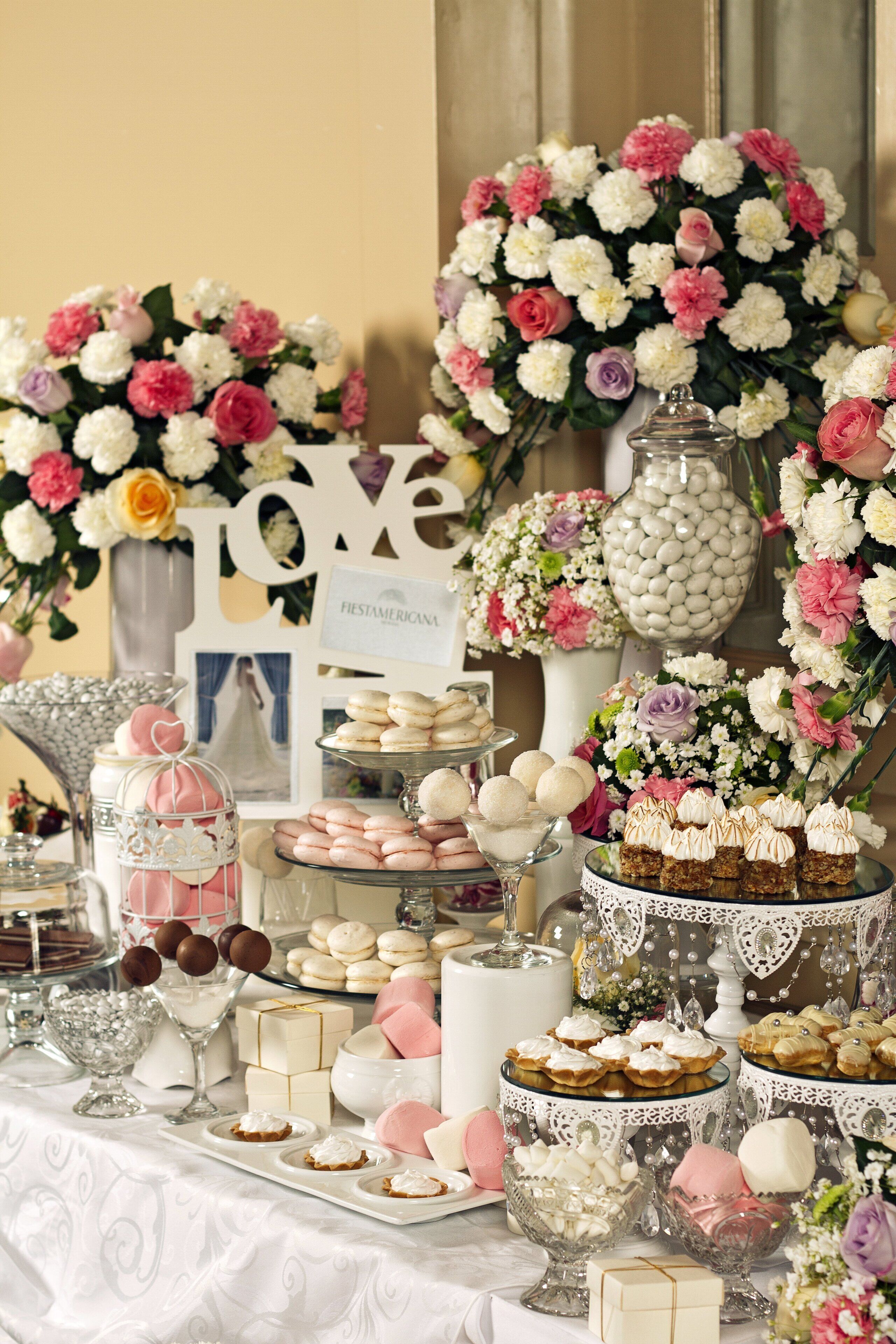 Uma mesa com diversos doces e arranjos florais, incluindo uma placa com a inscrição "FIESTA AMERICANA".