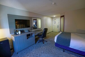Quarto de hotel com cama, escrivaninha com cadeira, televisão e pequeno frigorífico.