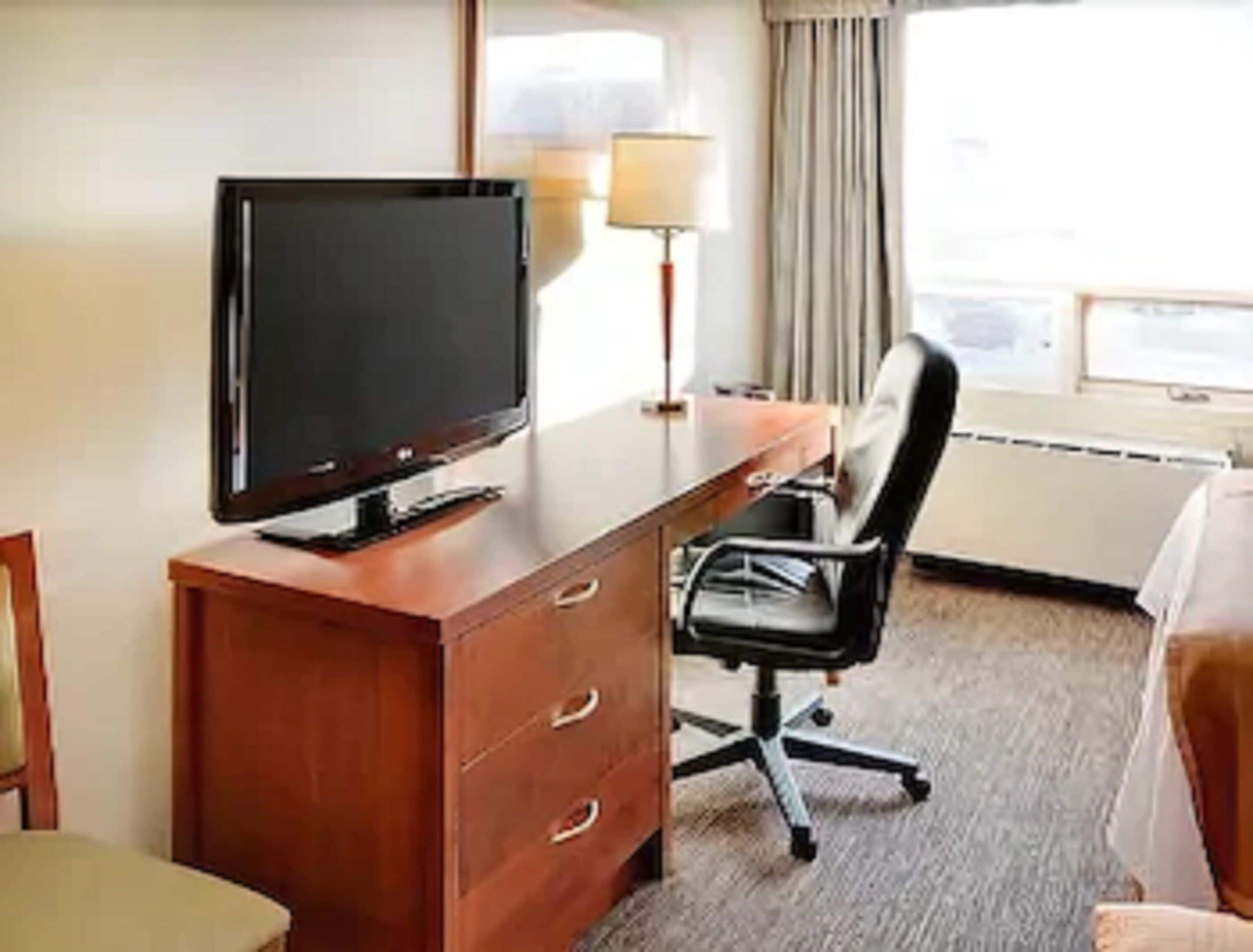 Quarto de hotel com secretária de madeira, televisão, cadeira de escritório preta e janela com cortinas.