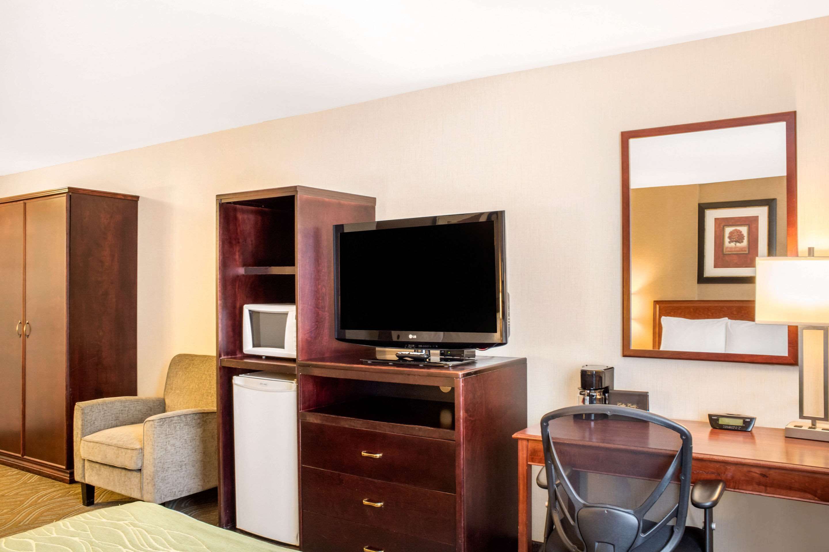 Quarto de hotel com cama, escrivaninha com cadeira, televisão, guarda-roupa e um pequeno frigorífico.