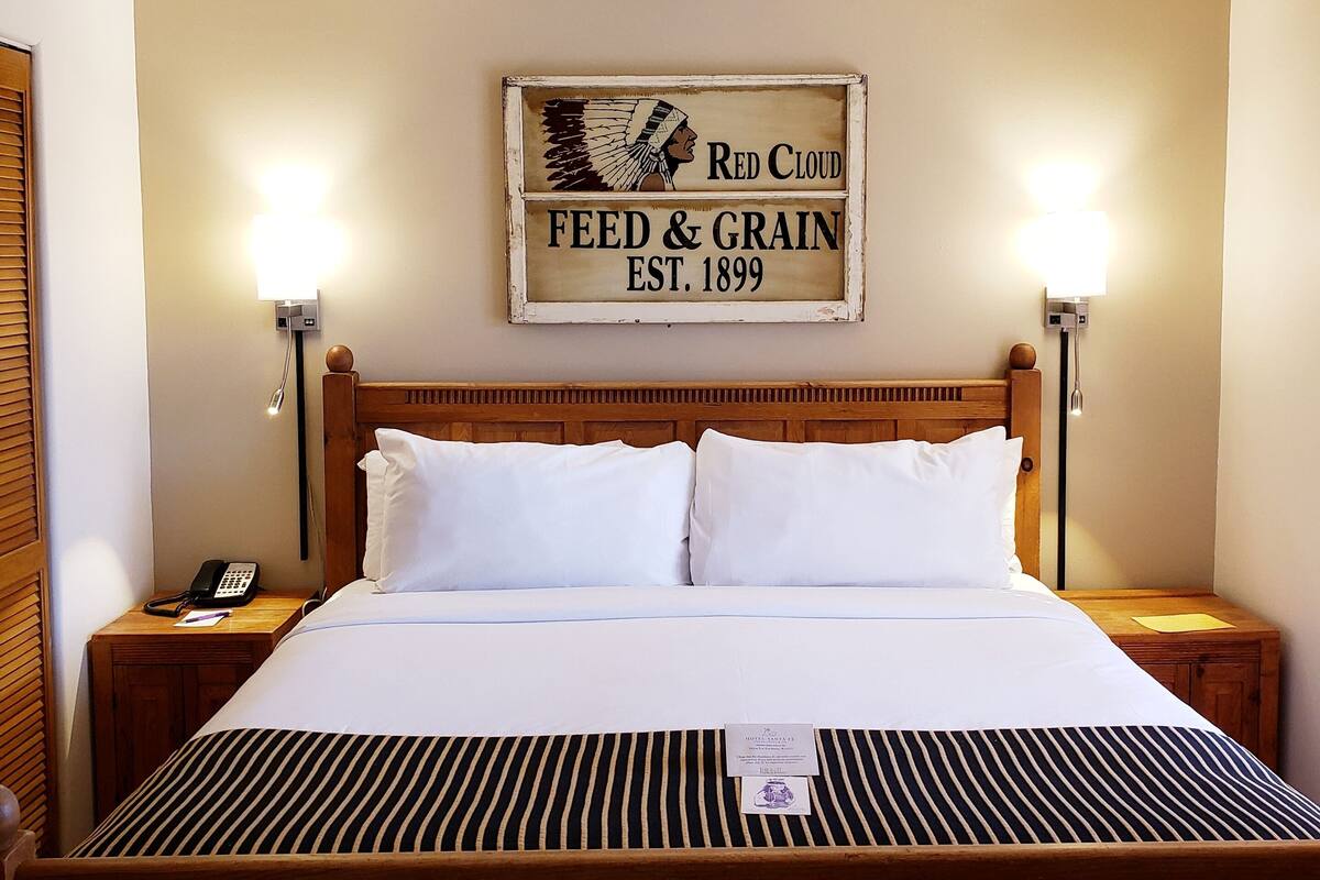 Quarto de hotel com cama de madeira, roupa de cama branca, duas luminárias de cabeceira, uma placa na parede e um telefone sobre uma mesa de cabeceira de madeira.
