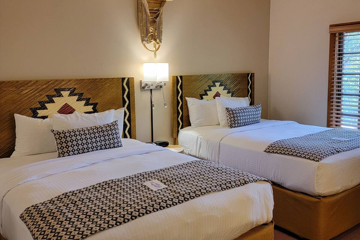 Quarto de hotel com duas camas, cada uma com roupa de cama e almofadas estampadas. Há uma luminária fixada na parede sobre as camas e uma janela com persianas à direita.