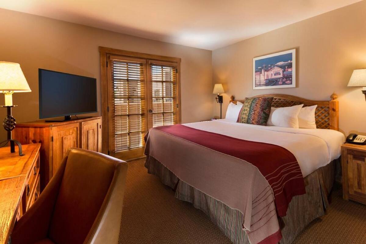 Quarto de hotel com cama, escrivaninha, televisão e janela com persianas.