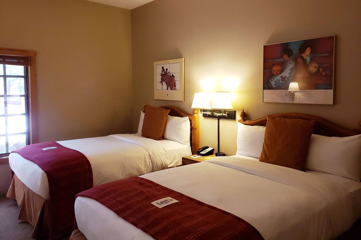 Quarto de hotel com duas camas, mesa de cabeceira com abajur e obras de arte nas paredes.