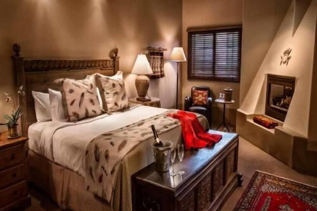 Quarto com cama de madeira, mesa de cabeceira, abajur, janela com persianas e tapete vermelho.