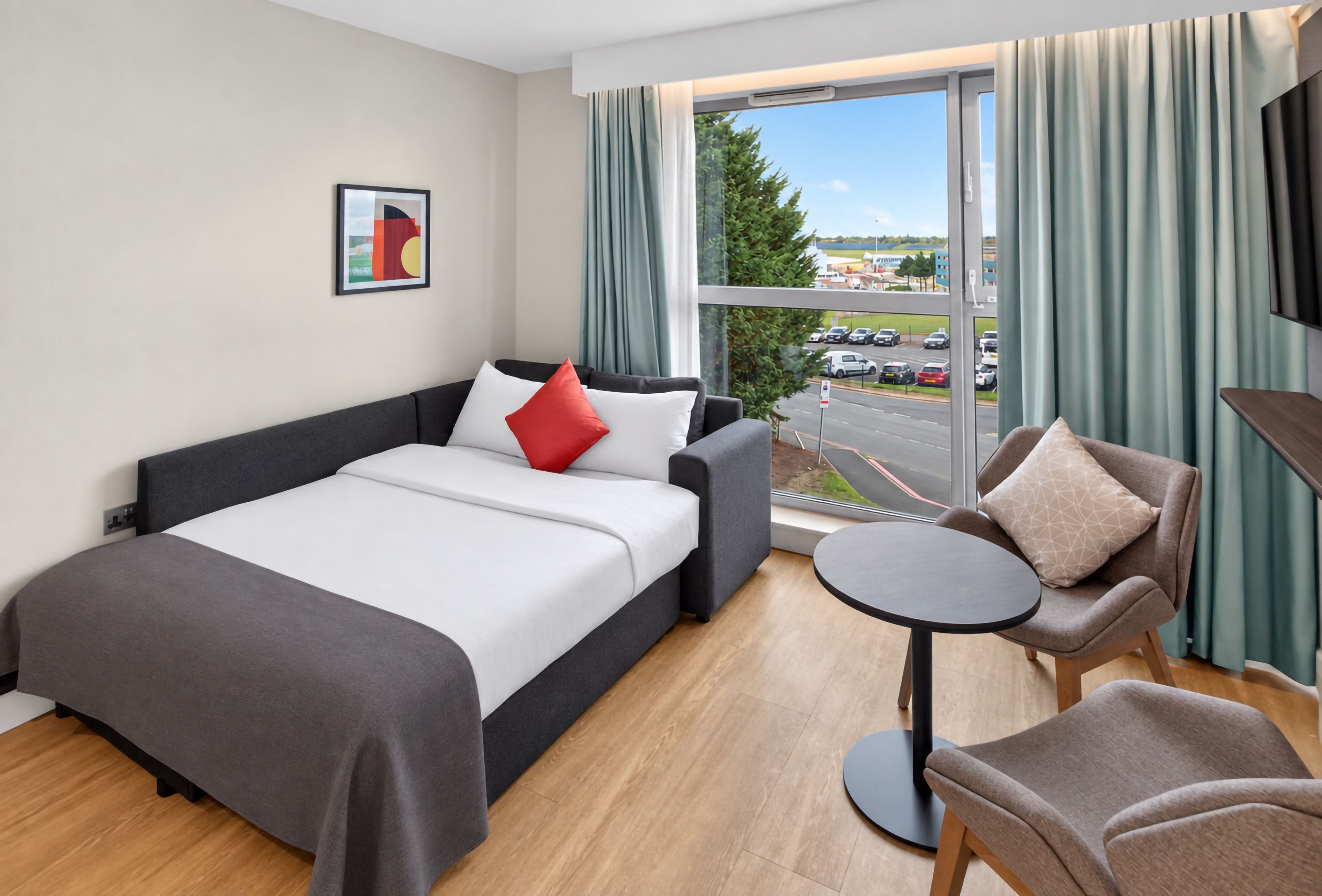 Quarto de hotel com cama, sofá, mesinha e vista para um estacionamento pela janela.