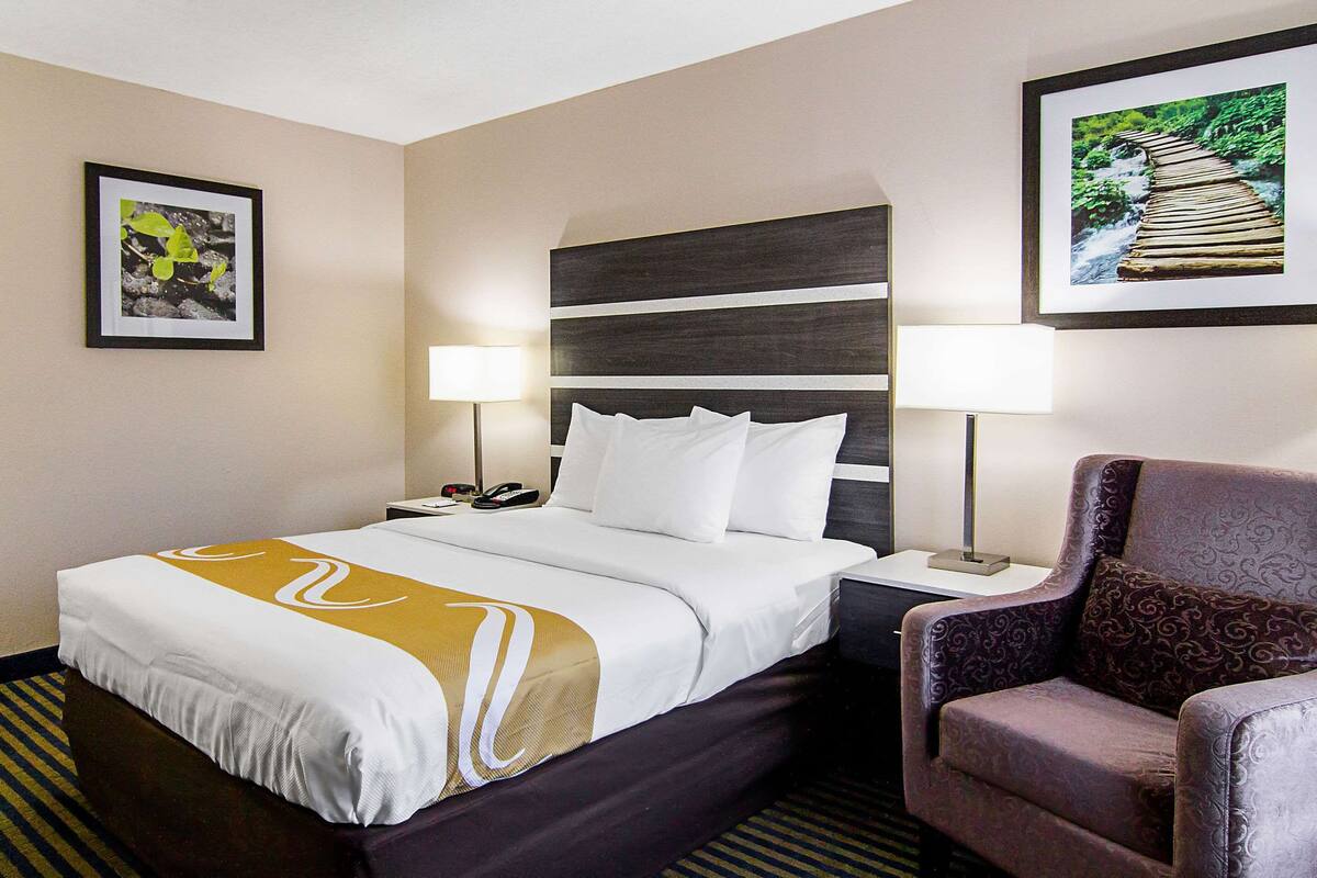 Quarto de hotel com uma cama grande, duas mesas de cabeceira com luminárias, uma cadeira e quadros nas paredes.
