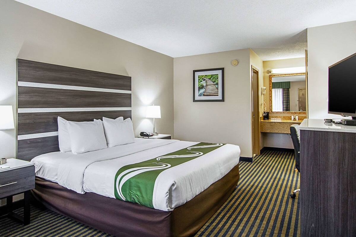 Quarto de hotel com cama, mesa de cabeceira, luminária, escrivaninha, cadeira e televisão.