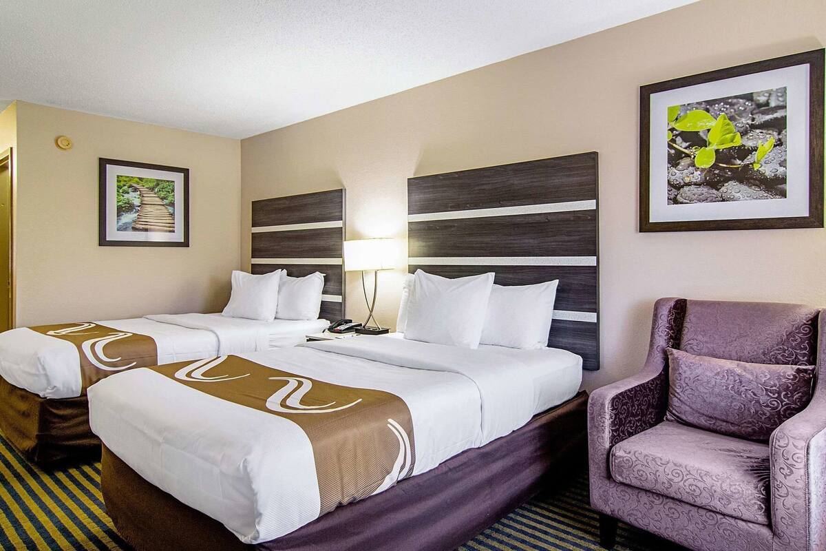 Quarto de hotel com duas camas, uma mesa de cabeceira, uma luminária, uma cadeira e duas imagens emolduradas nas paredes.