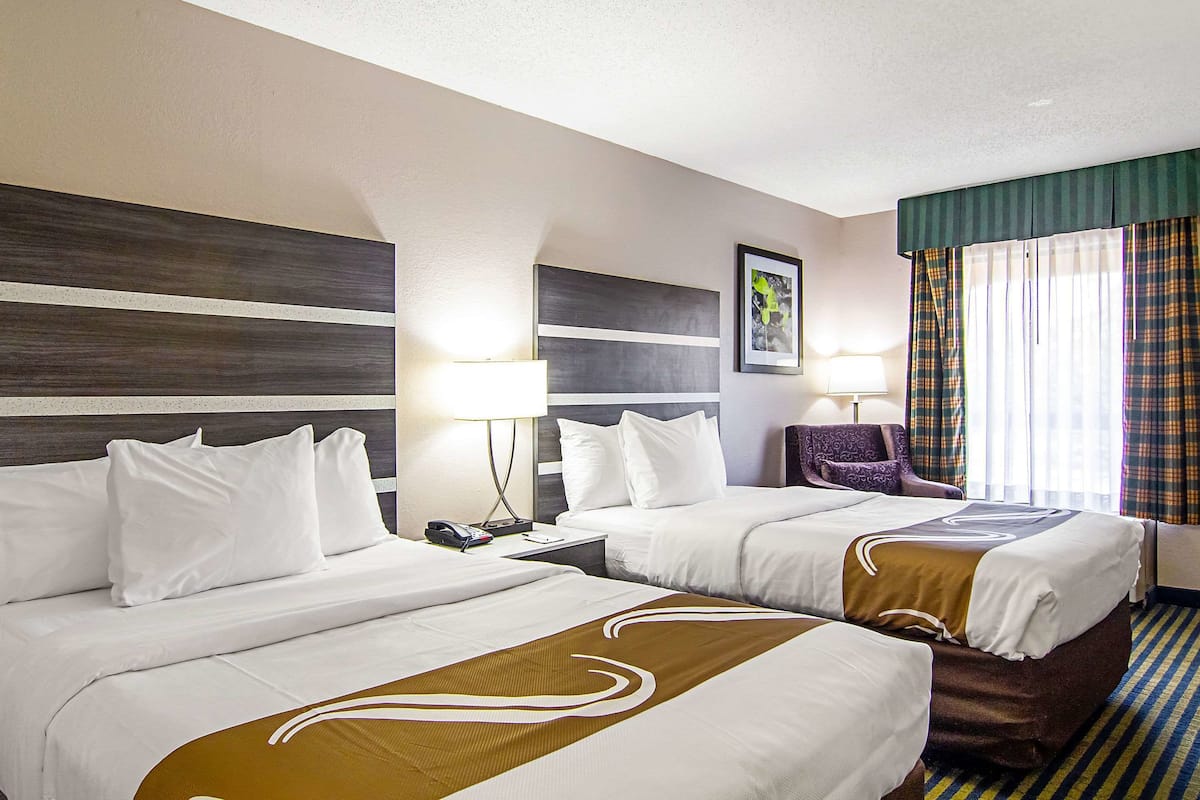 Quarto de hotel com duas camas, mesa de cabeceira com abajur, uma cadeira e janela com cortinas.