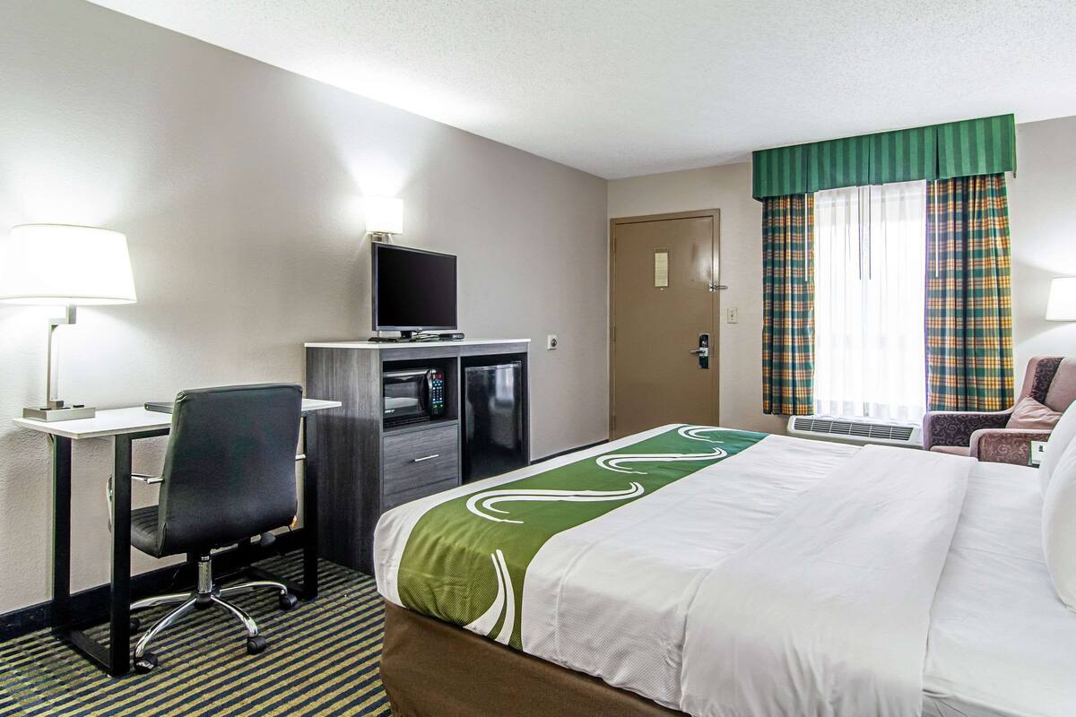 Quarto de hotel com cama, escrivaninha com computador, cadeira, televisão e porta.