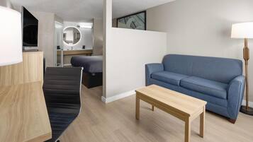 Quarto de hotel moderno com sofá, mesa de madeira, cadeira preta, TV, espelho e abajur.