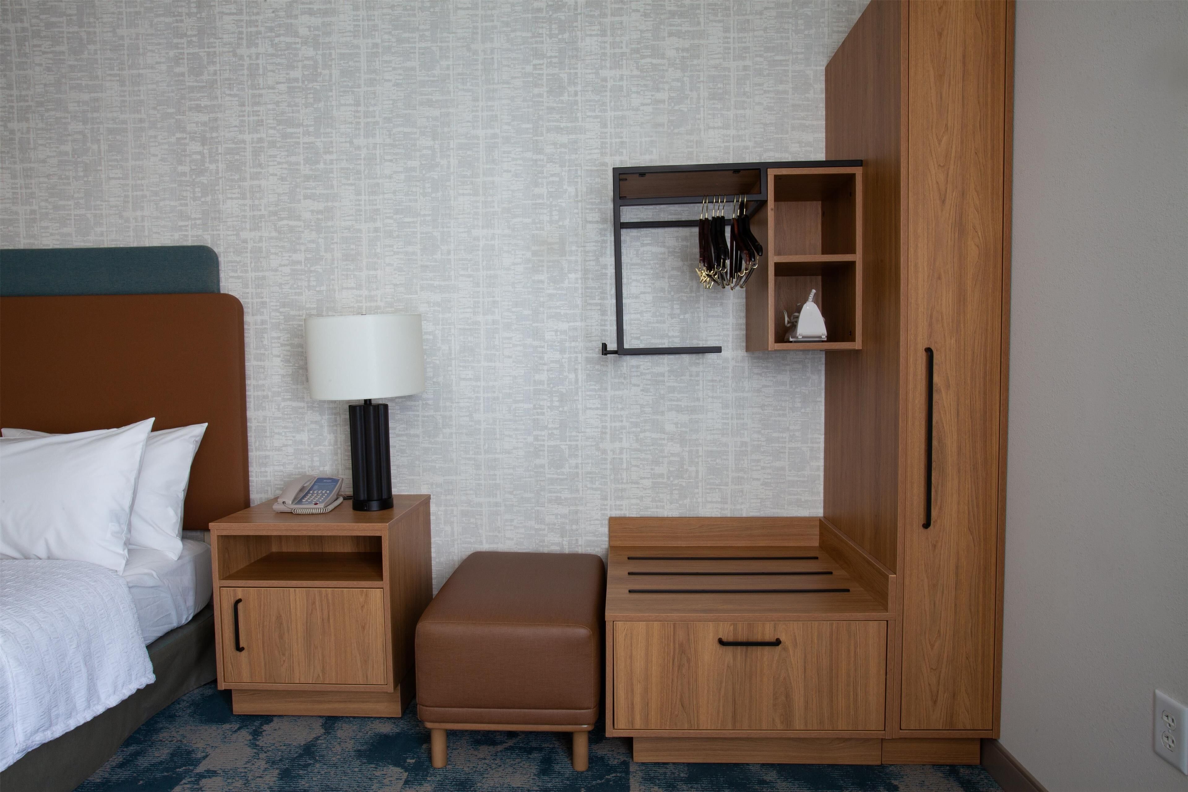 Quarto de hotel com cama, mesa de cabeceira com abajur, criado-mudo de madeira, guarda-roupa de madeira e um pequeno armário de madeira.