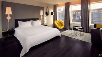 Quarto de hotel moderno com uma cama grande, uma poltrona amarela, uma mesa pequena e vista para a cidade através de amplas janelas.