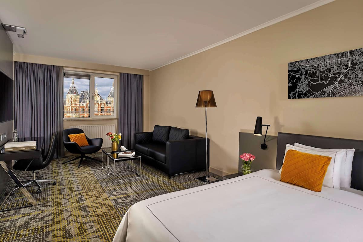 Quarto de hotel com cama, escrivaninha, cadeira, sofá, luminária e vista para a cidade através de grandes janelas.