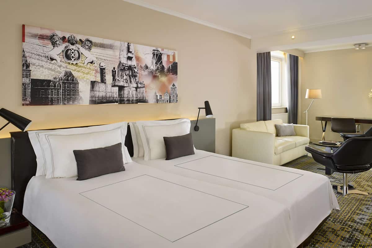 Quarto de hotel com uma cama grande, uma escrivaninha, uma cadeira e um sofá. Há um quadro na parede e um vaso com flores sobre a escrivaninha.