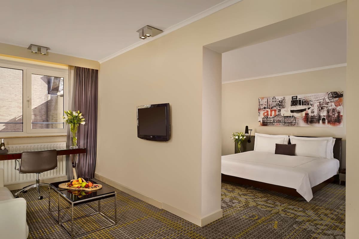 Quarto de hotel com cama, escrivaninha com cadeira, mesinha com frutas, janela com cortinas e televisão fixada na parede.