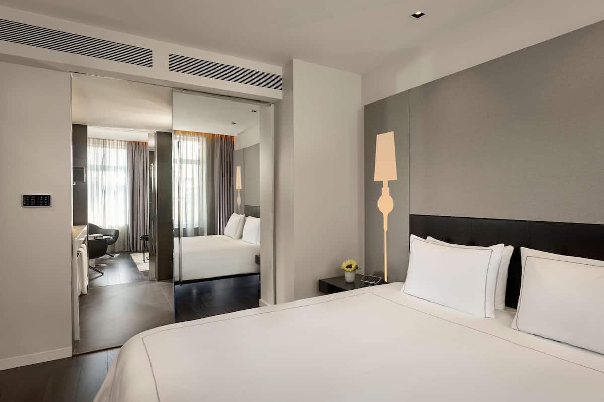 Quarto de hotel moderno com uma cama grande, uma mesa de cabeceira com luminária, um espelho e vista para o corredor e outro quarto.