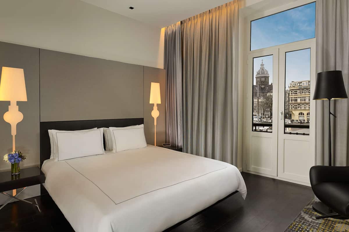 Quarto de hotel com uma cama grande, duas luminárias de cabeceira, uma cadeira e vista para a paisagem urbana pela janela.