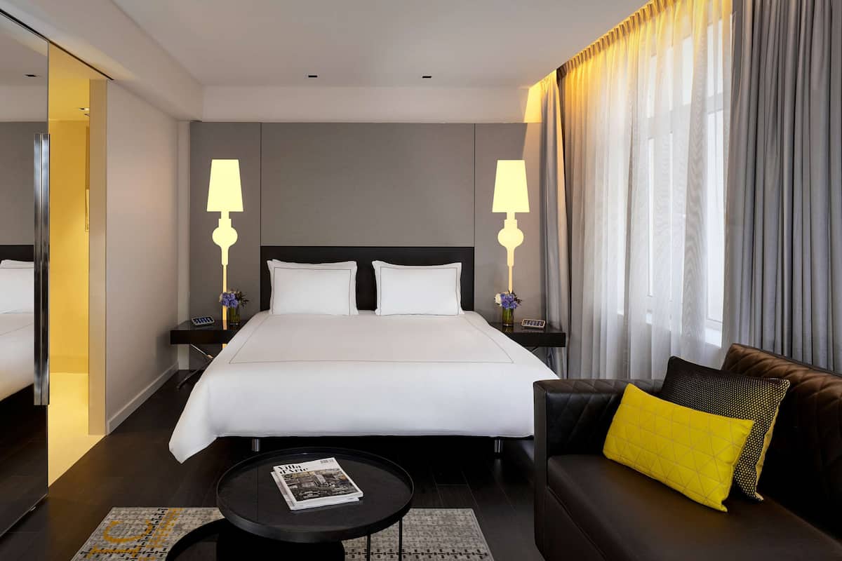 Quarto de hotel moderno com uma cama grande, duas mesinhas de cabeceira com luminárias, um sofá com um travesseiro amarelo e uma pequena mesa de centro.