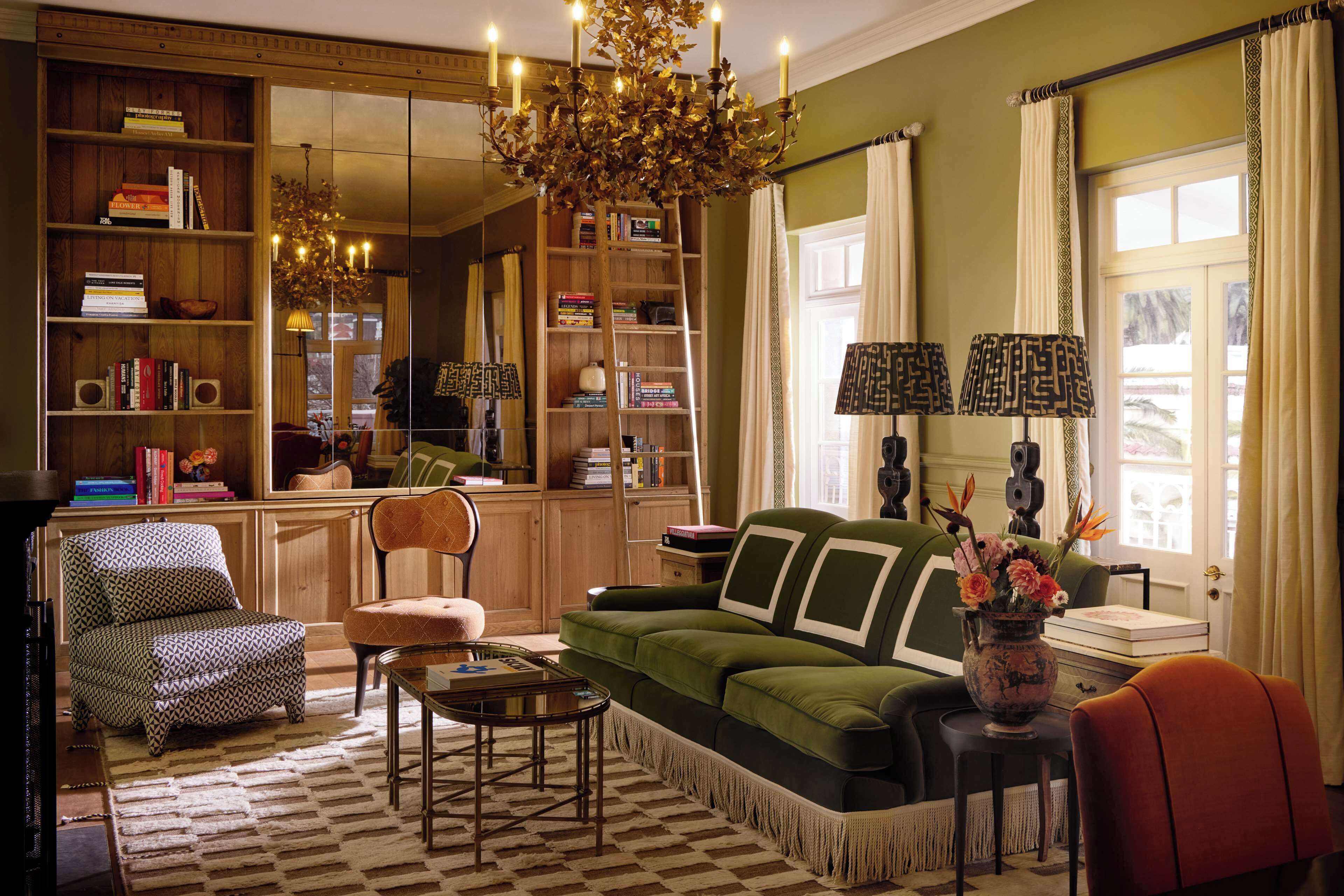 Uma sala de estar aconchegante com um sofá verde, poltronas estampadas, uma estante de madeira e um lustre decorativo.