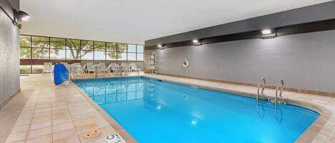 Piscina interna com água cristalina, piso ladrilhado e vista para árvores através de grandes janelas.