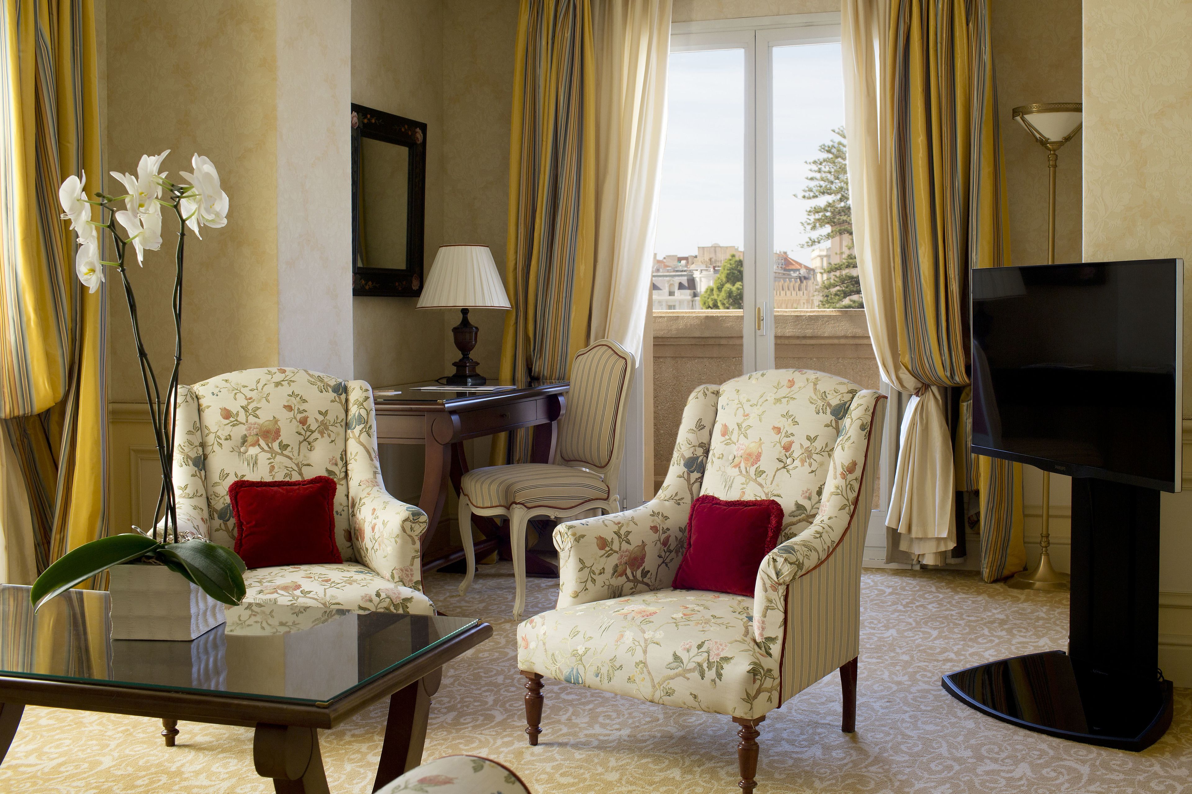 Quarto de hotel com duas poltronas florais, uma mesa de centro de vidro, uma escrivaninha com luminária, uma TV de tela plana e vista para a cidade pela janela.