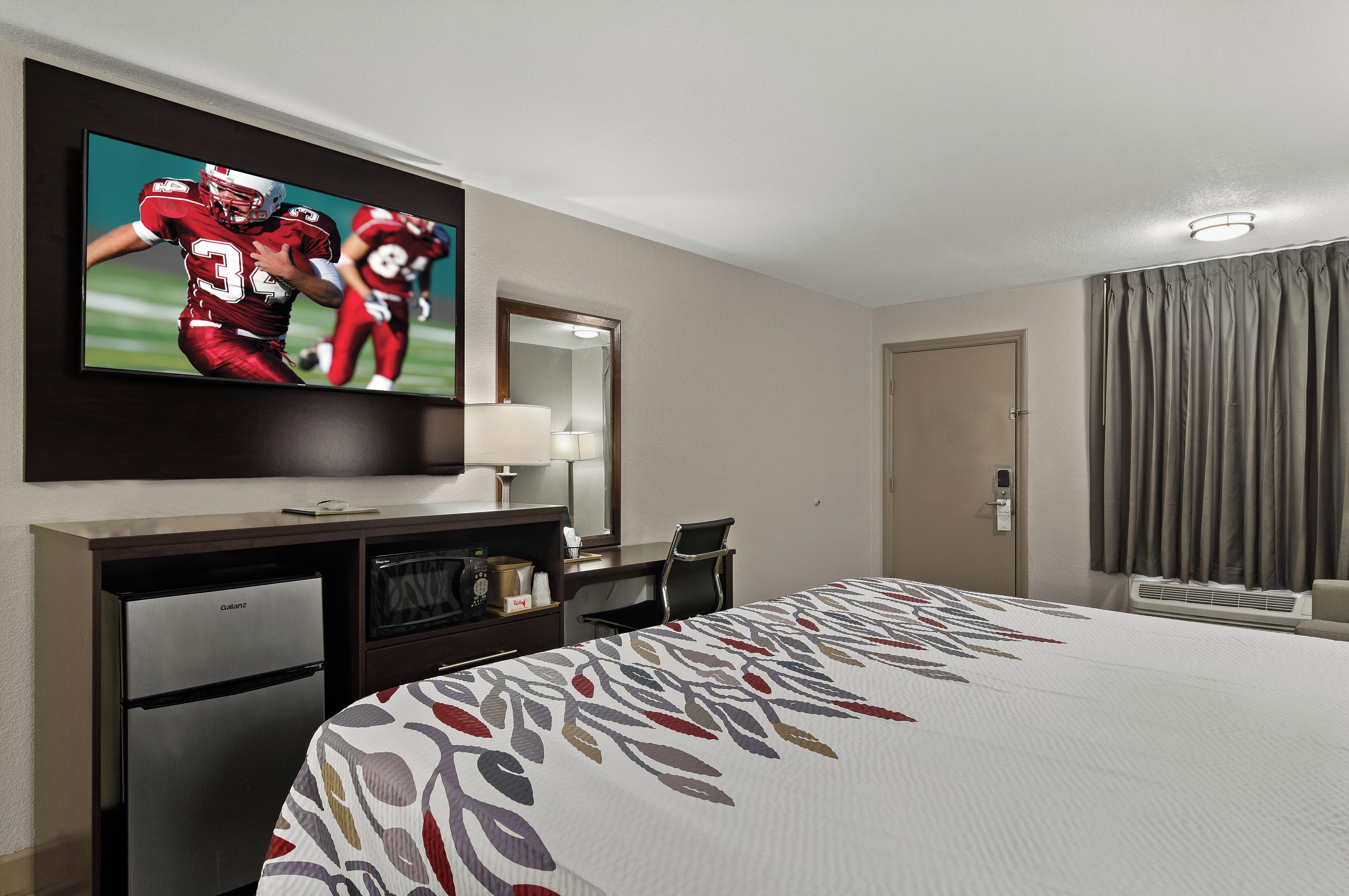Quarto de hotel com uma cama grande, televisão de tela plana na parede, escrivaninha com computador, mini-geladeira e luminária.
