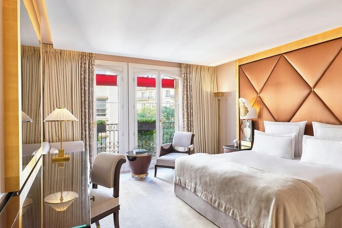 Quarto de hotel com cama, duas cadeiras, uma mesinha, luminária e vista exterior.