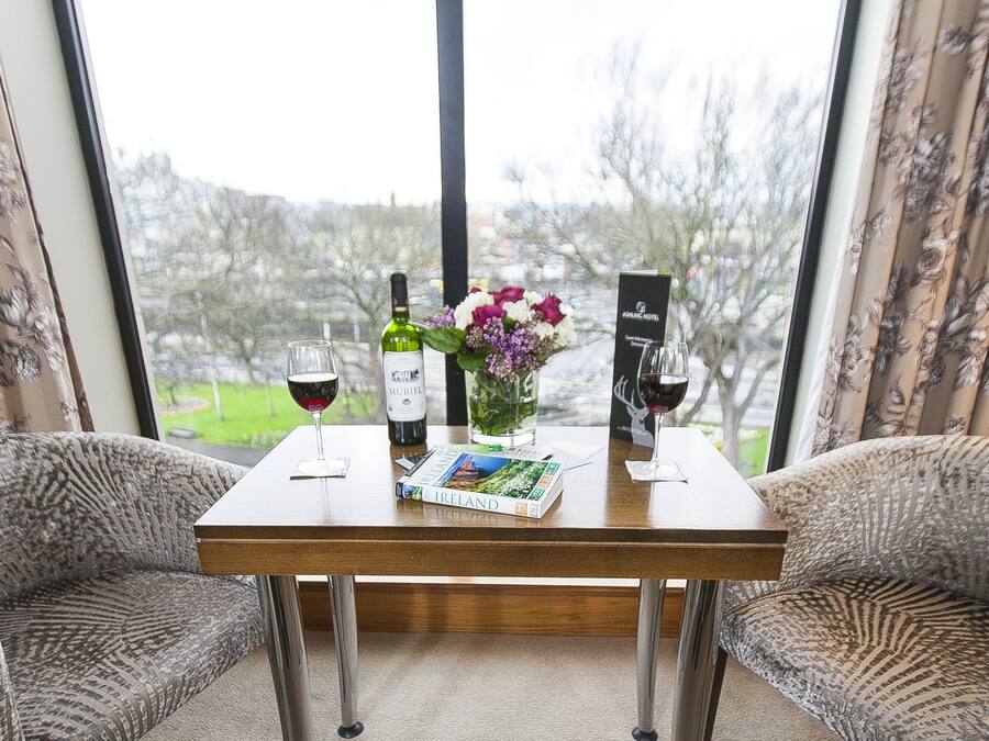 Quarto com uma janela grande, duas poltronas estampadas, uma mesa de madeira com um livro, uma garrafa de vinho e uma taça de vinho.