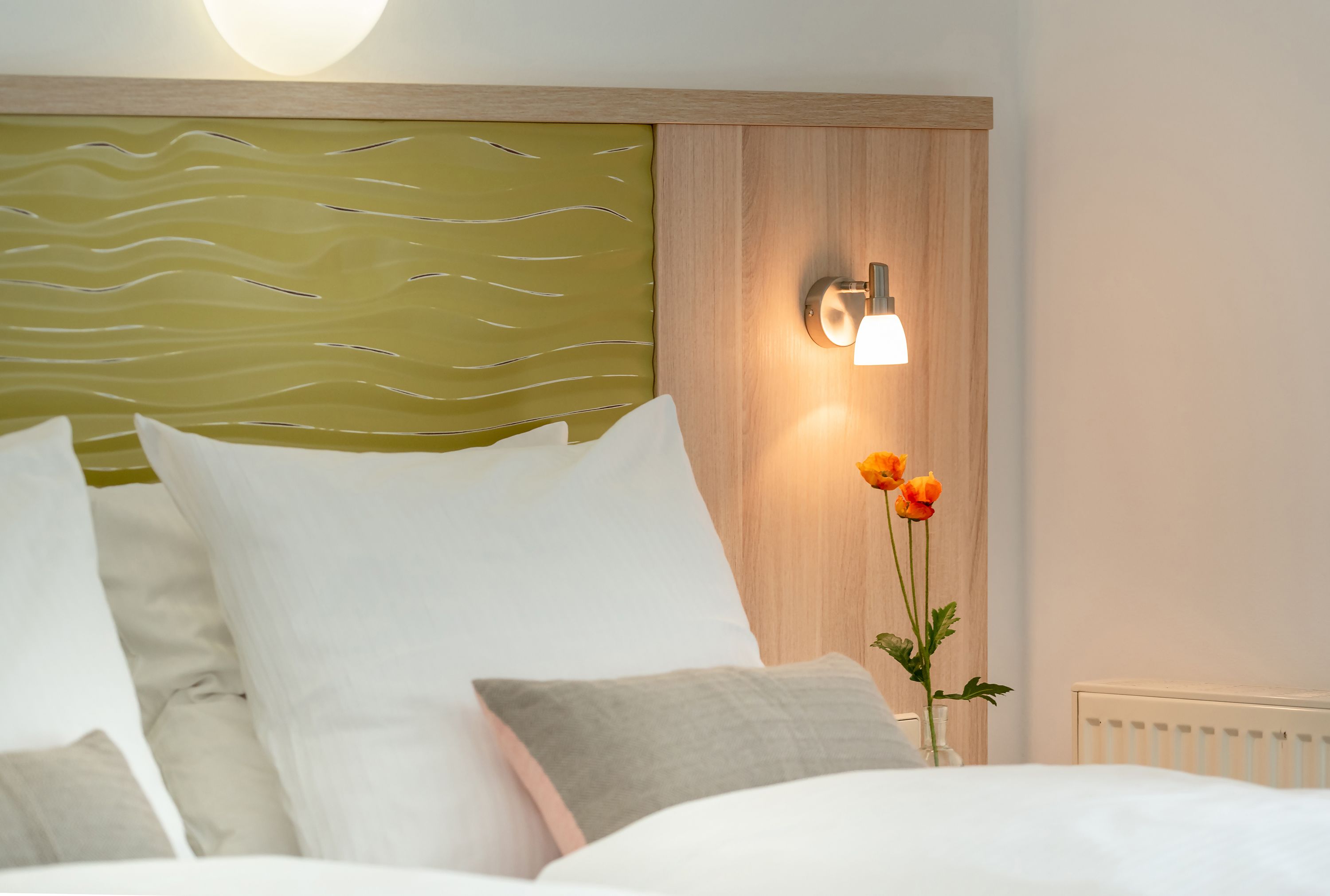 Quarto de hotel com cama, travesseiros, mesa de cabeceira, vaso com flores e luminária fixada na parede.