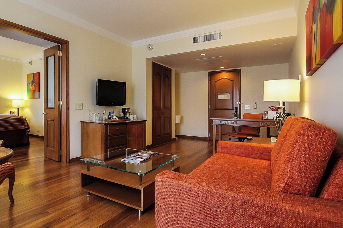 Quarto de hotel com área de estar, espaço para refeições, televisão e porta que dá para outro cômodo.