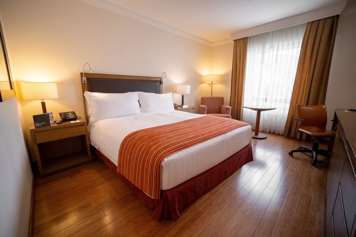Quarto de hotel com uma cama grande, mesas de cabeceira, escrivaninha, cadeira, abajur e janela com cortinas.