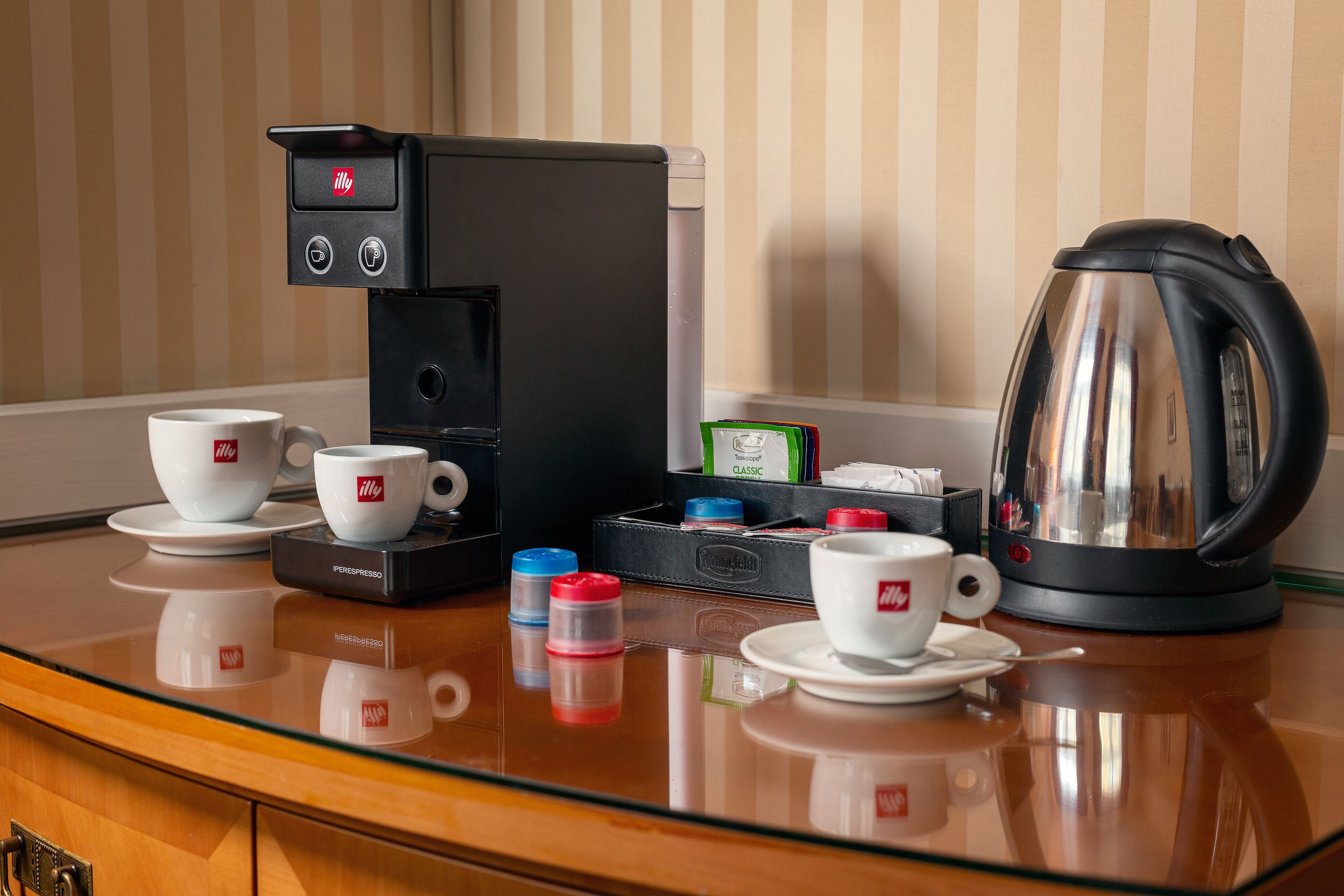 Uma estação de café com cafeteira, chávenas e chaleira sobre uma mesa de vidro.