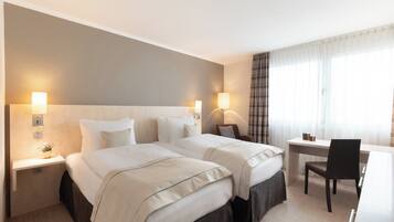 Quarto de hotel com duas camas, uma mesa de trabalho e uma cadeira. Há abajures e uma janela com cortinas.