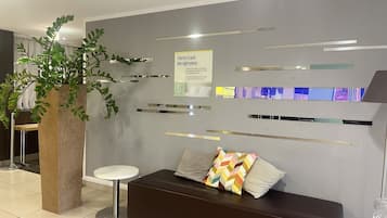 Um lobby de hotel moderno com uma receção, uma grande planta em vaso, um sofá com almofadas coloridas e um cartaz de mensagens inspiradoras na parede.