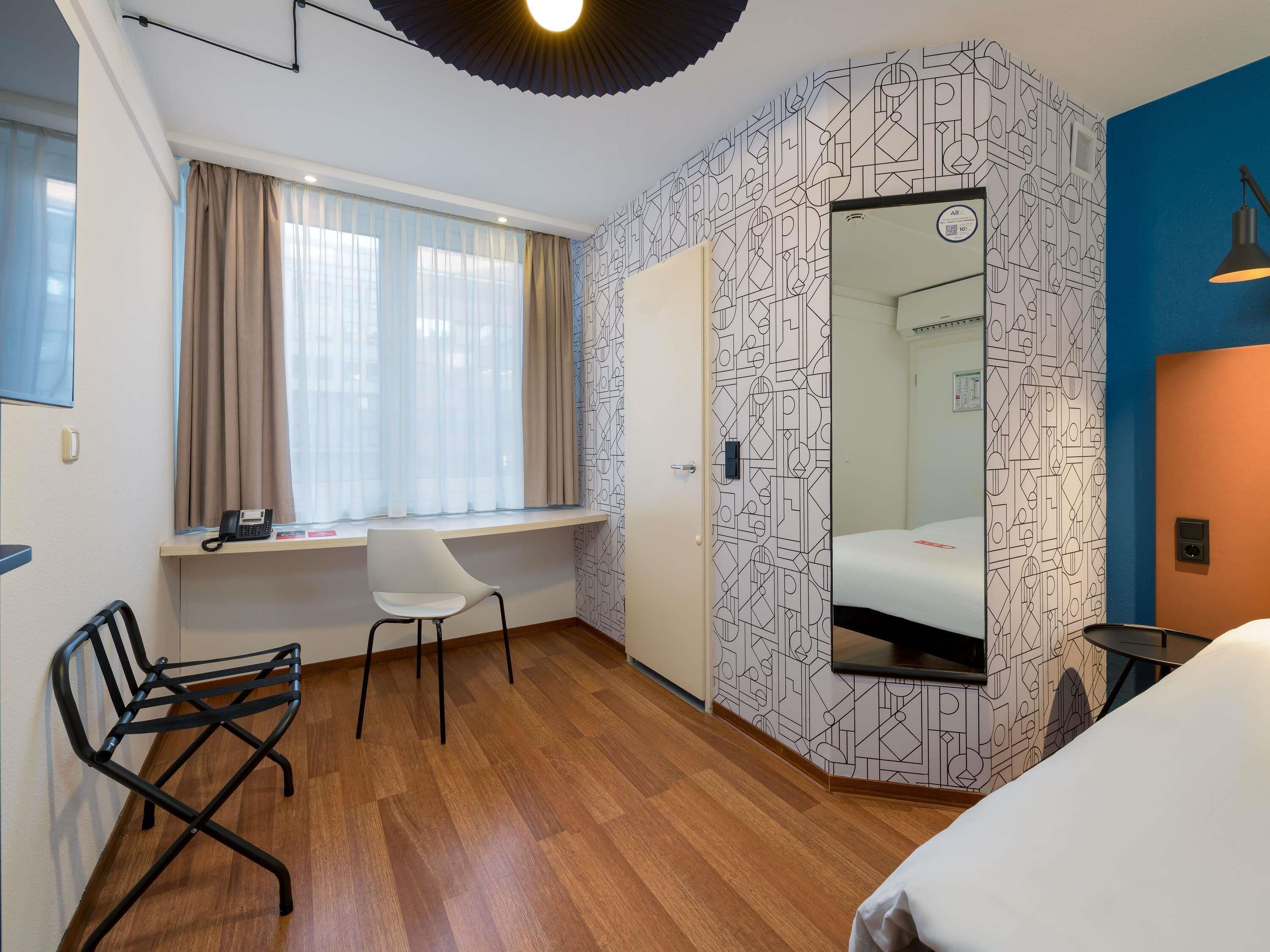 Quarto de hotel com piso de madeira, uma escrivaninha com cadeira, uma cama, um espelho grande e uma janela com cortinas.