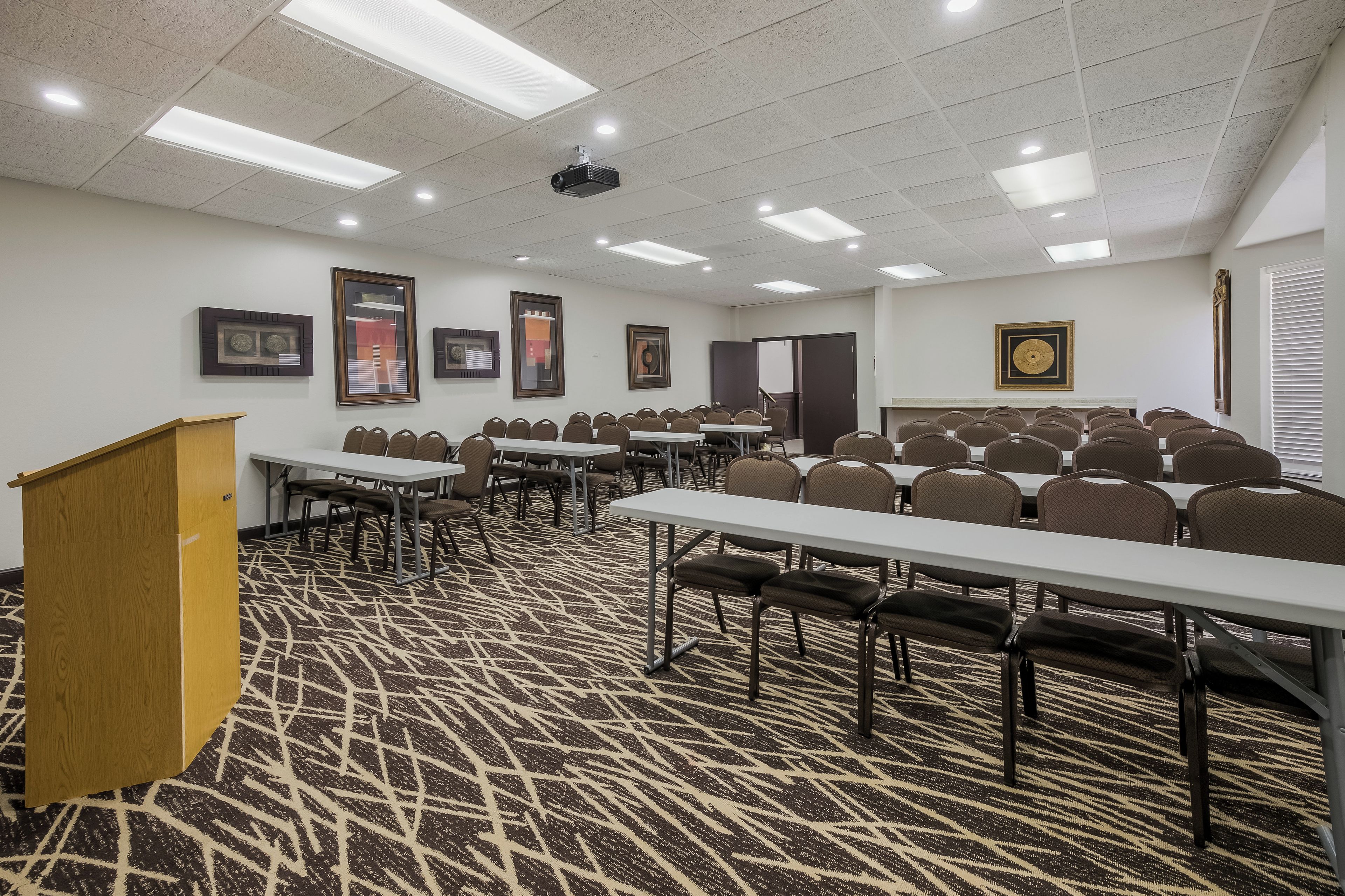 Sala de conferências com várias mesas e cadeiras, piso coberto de carpete e iluminação no teto.