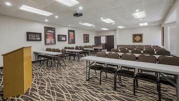 Sala de conferências com várias mesas e cadeiras, piso coberto de carpete e iluminação no teto.