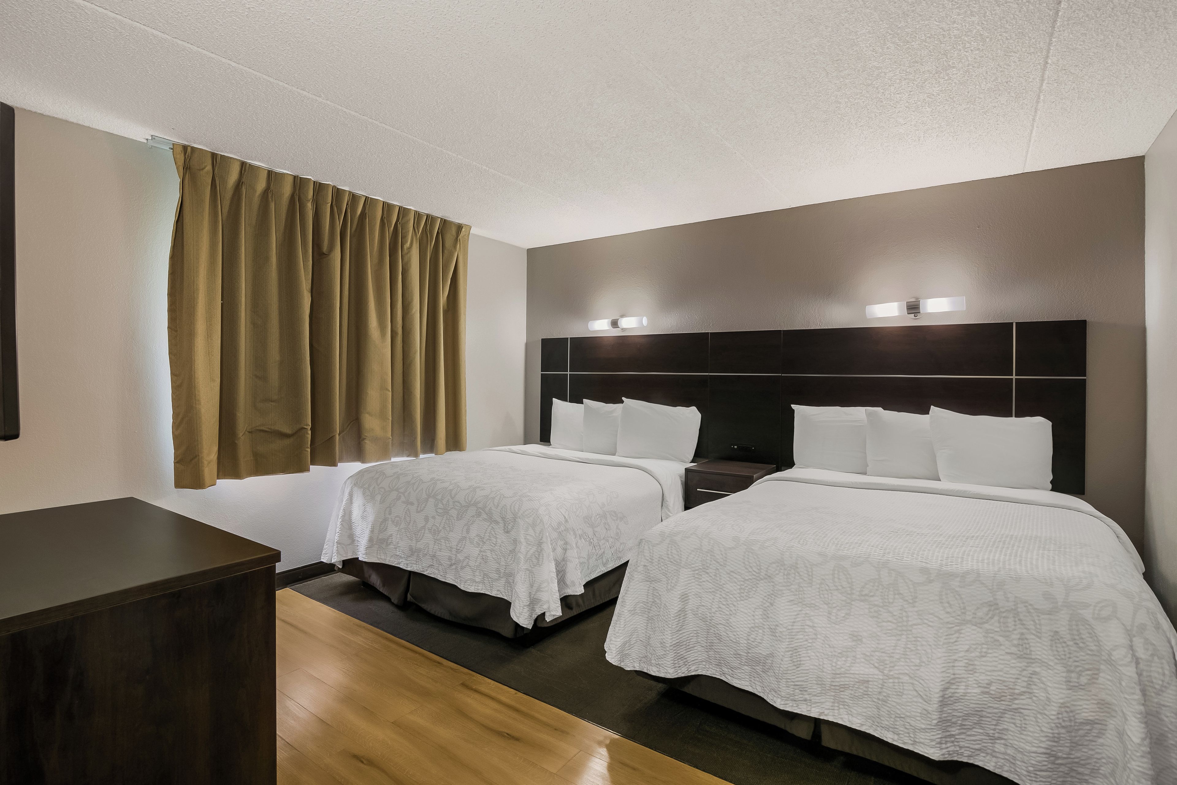 Quarto de hotel com duas camas, cômoda de madeira, janela com cortinas e luminária na parede.
