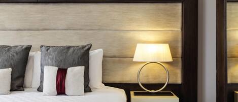 Cama bem arrumada com lençóis brancos e um cobertor vermelho, com almofadas cinzas e vermelhas de cada lado. Uma luminária de cabeceira com cúpula branca e base metálica está sobre uma mesa de cabeceira de madeira escura.