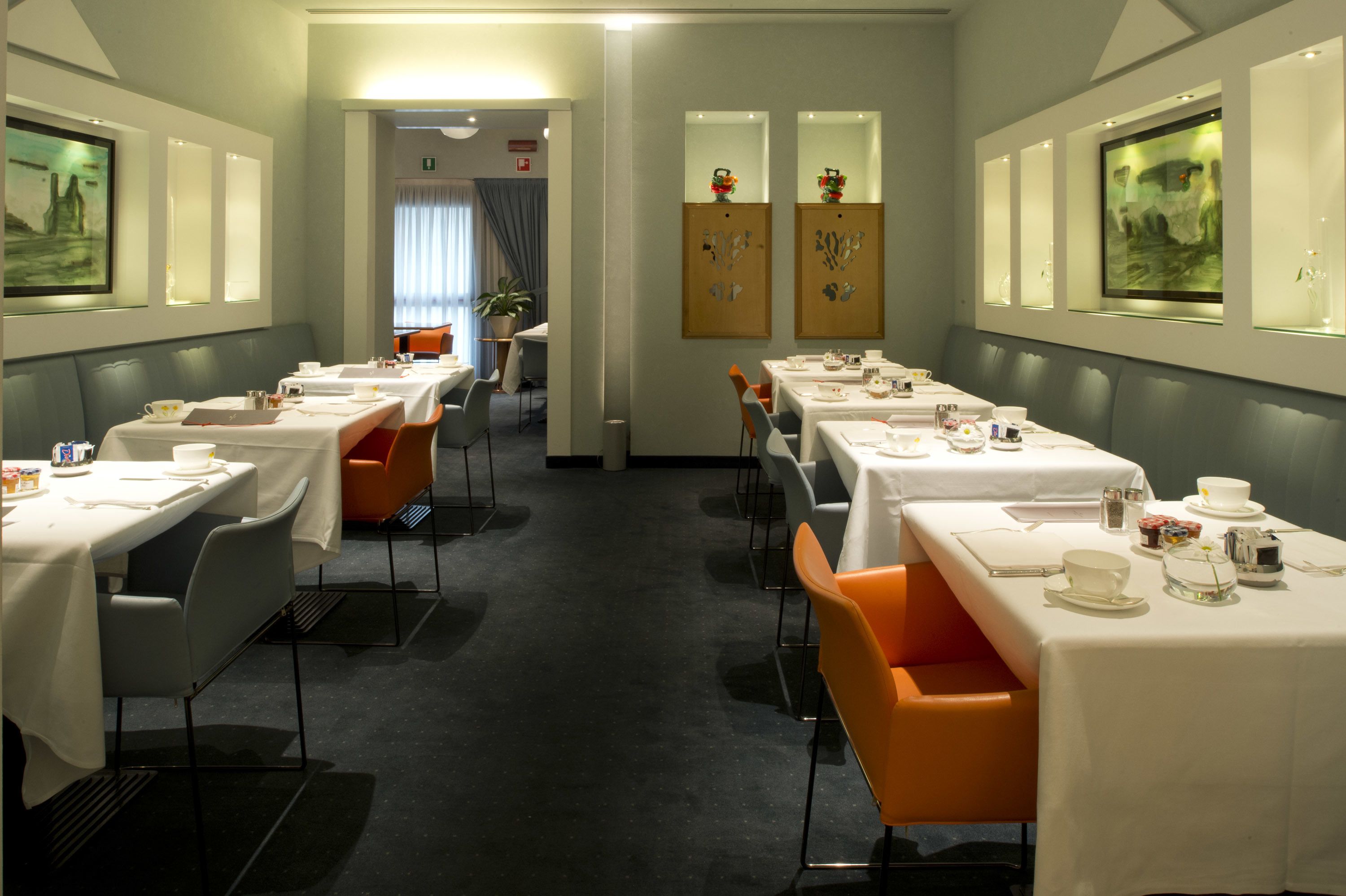 Uma sala de jantar de hotel com várias mesas preparadas para refeições, cadeiras e uma planta em vaso ao fundo.