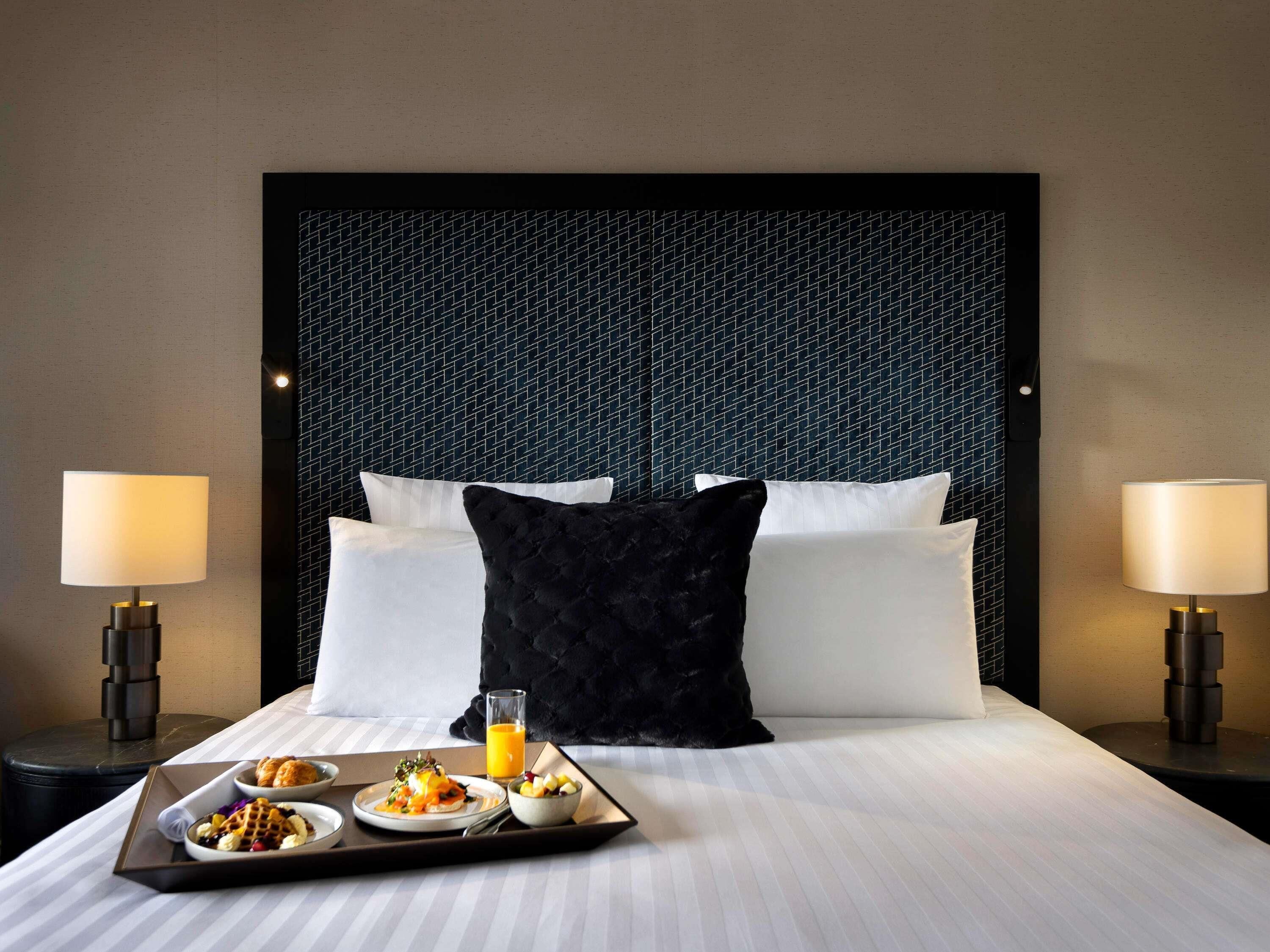 Quarto de hotel com uma cama grande, duas luminárias de cabeceira, um tabuleiro com café da manhã e um painel de cabeceira estampado.